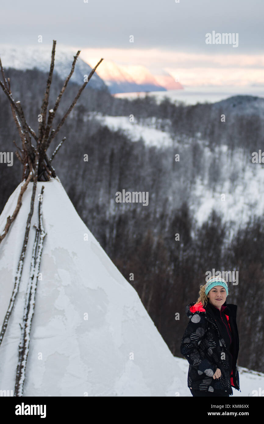 Girl smiles stood next to Sami lavvu as the sun stes on the Lyngen alps ...