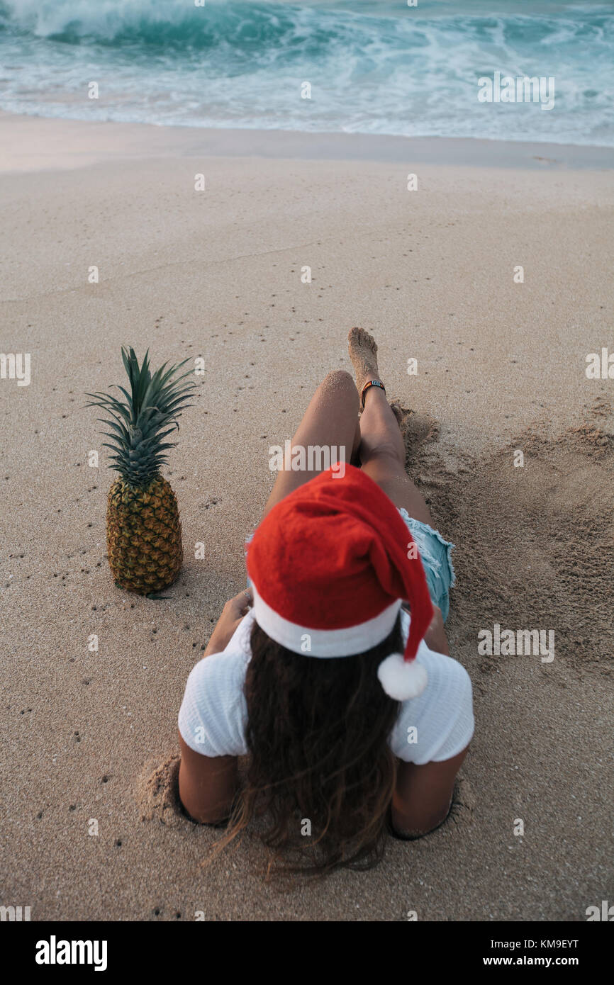 Woman Wearing A Christmas Santa Hat Lying On Beach Next To A Pineapple