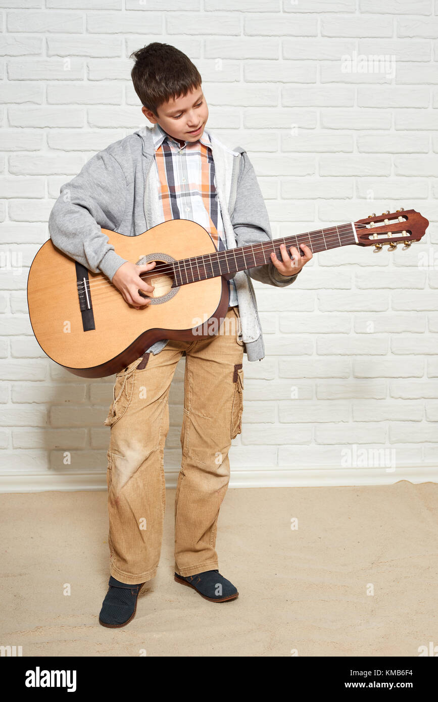 boy playing music on guitar, standing at full height, white brick wall ...