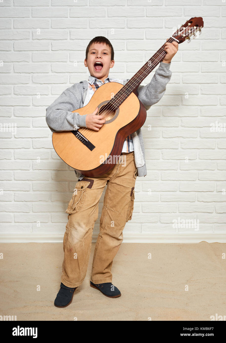 boy playing music on guitar, standing at full height, white brick wall ...