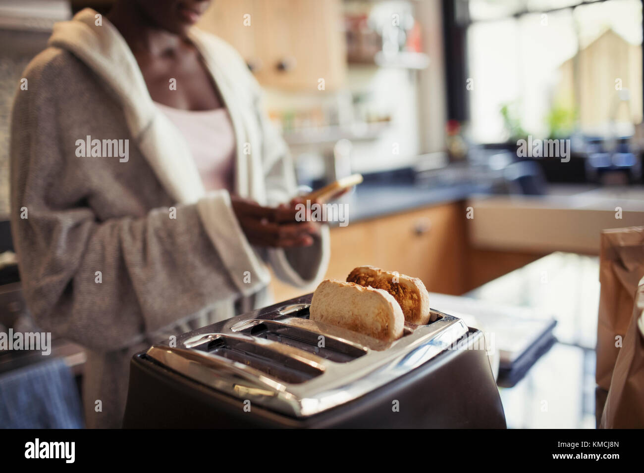 Woman texting with smart phone, toasting bread in toaster in kitchen ...