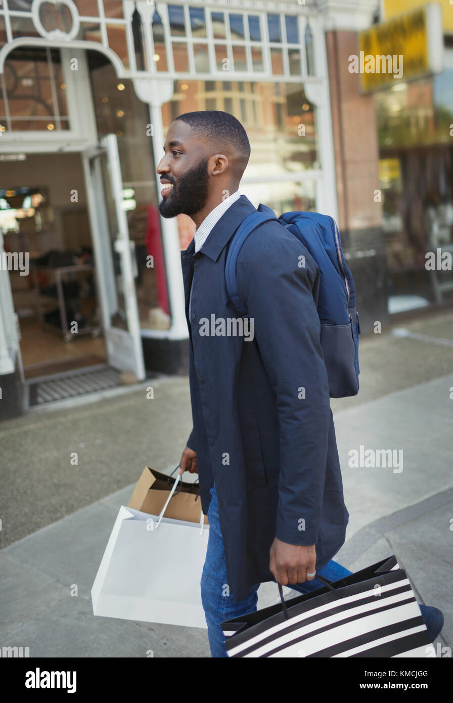 Smiling young man walking along storefront, carrying shopping bags ...