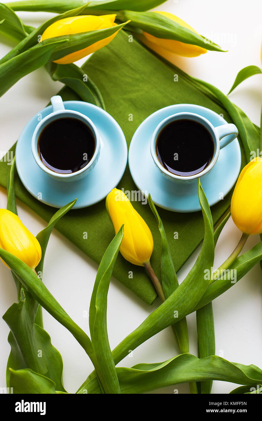 two blue cups of coffee on a table with green cloth and yellow tulips ...