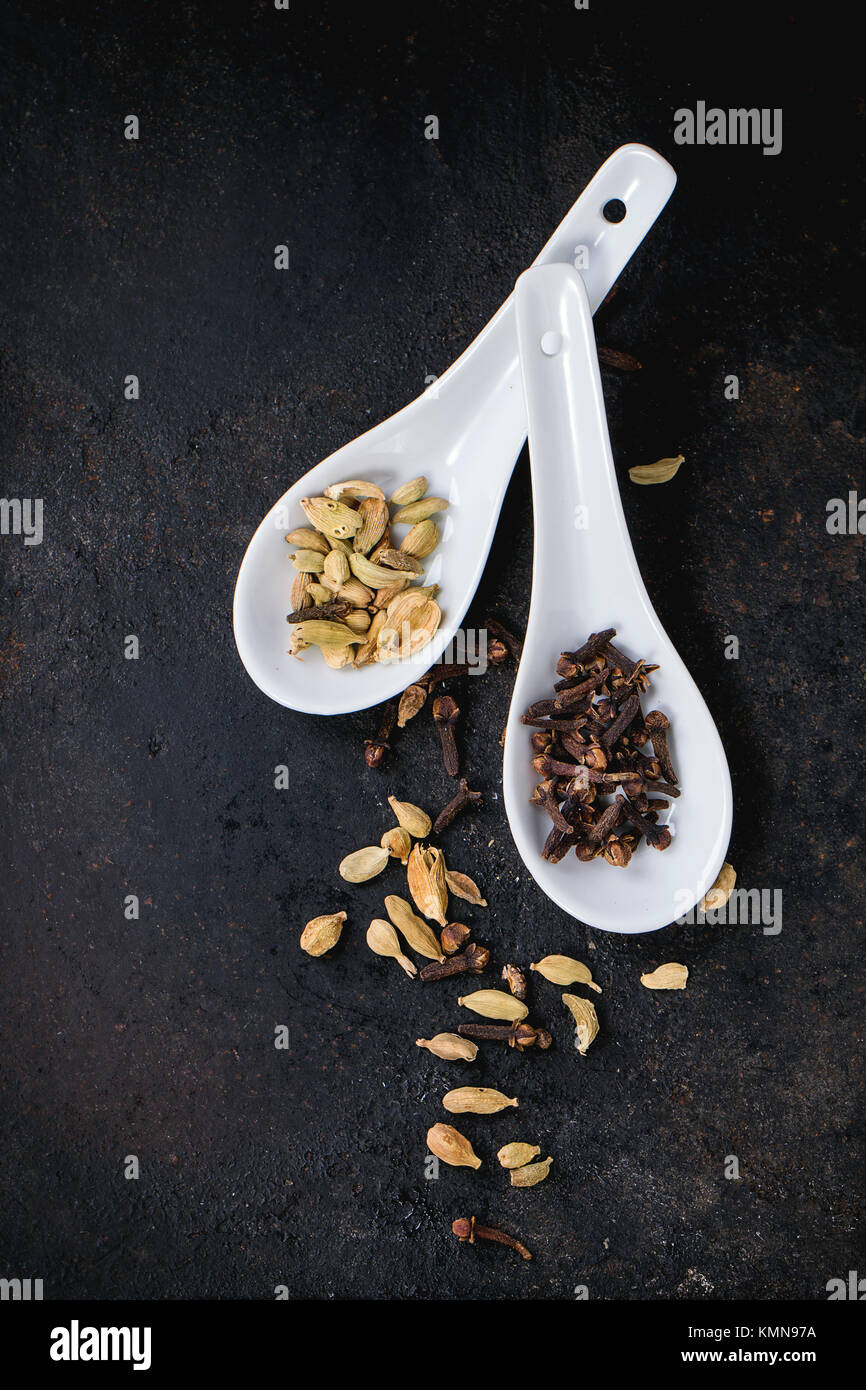 Cardamom seeds and cloves in white ceramic spoons over black background ...
