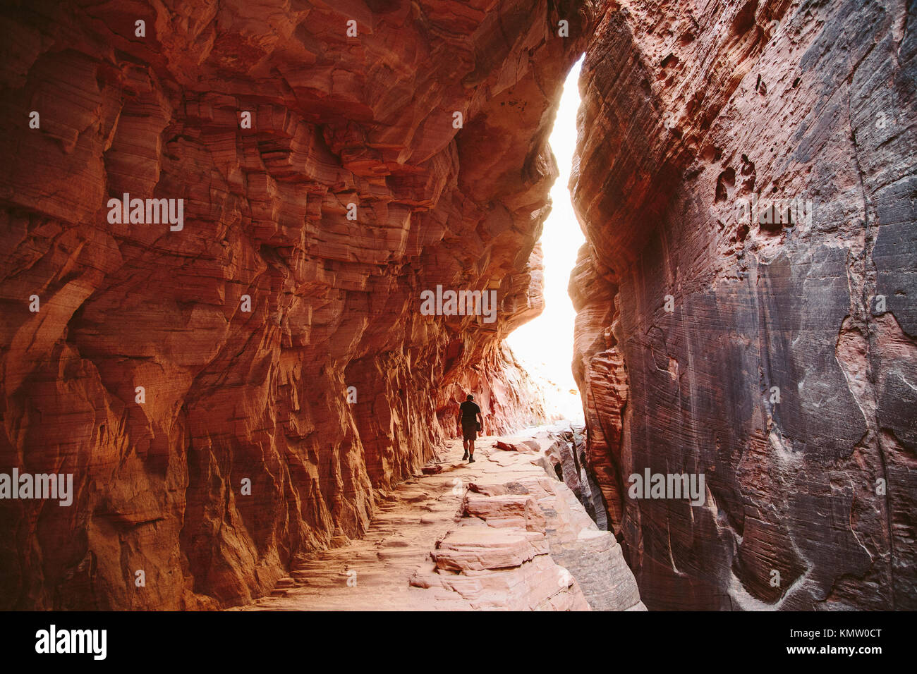 A hiker walks on a rock path inside a canyon with narrow red rock walls ...