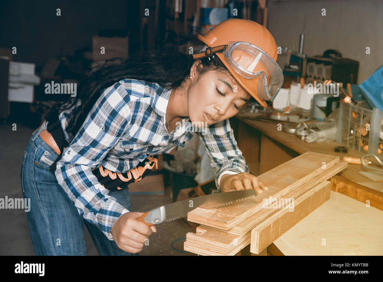 Pretty Asian woman using a hand saw to cut some wood in a wood shop ...