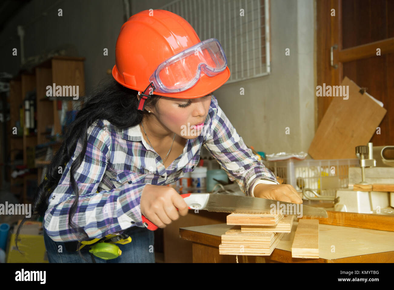 Pretty Asian woman using a hand saw to cut some wood in a wood shop ...