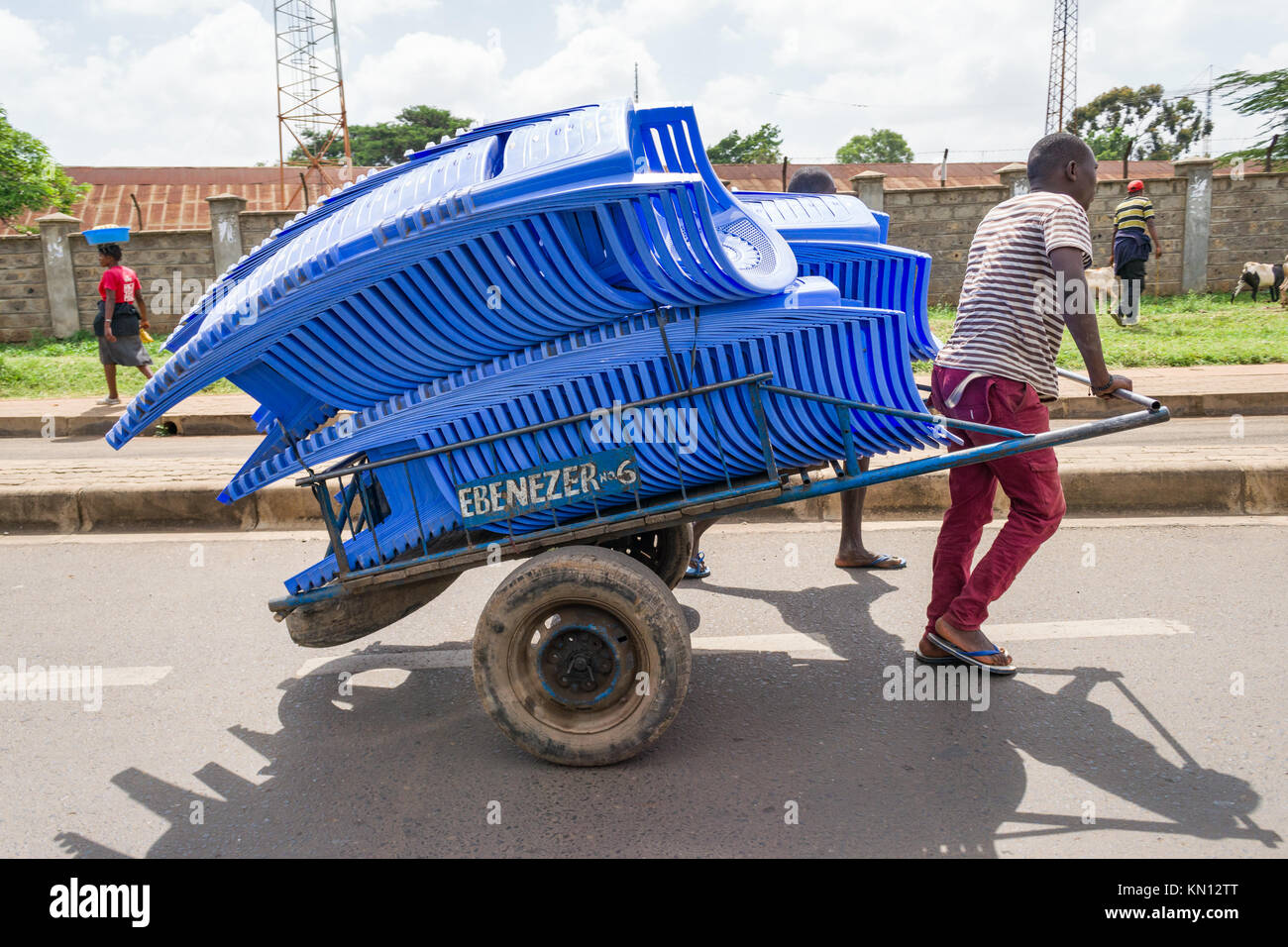 A Kenyan man pulling a handcart along a road carrying plastic chairs on ...