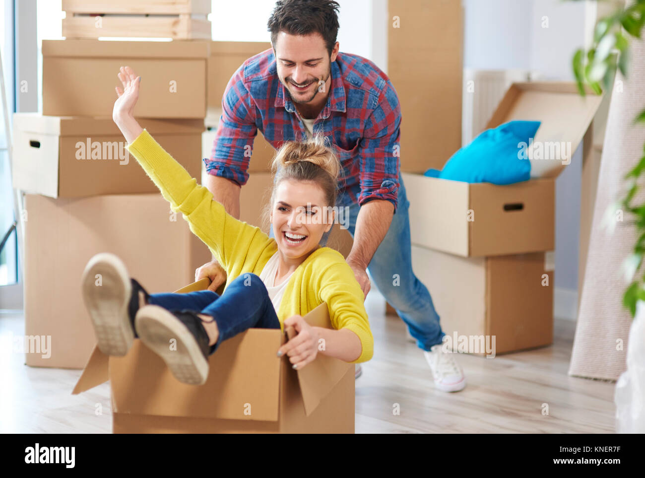 Young couple in new home, woman sitting in cardboard box, man pushing ...
