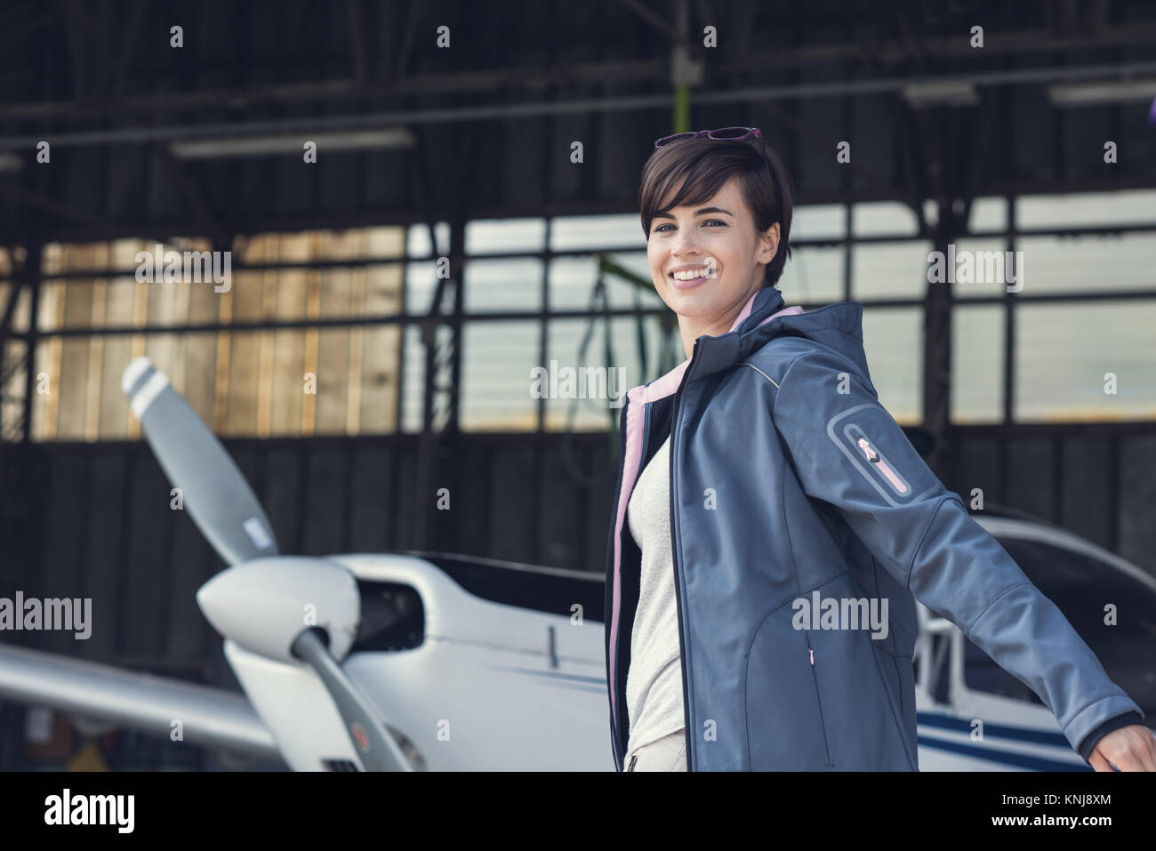 Smiling cheerful female pilot posing in the hangar with arms raised ...