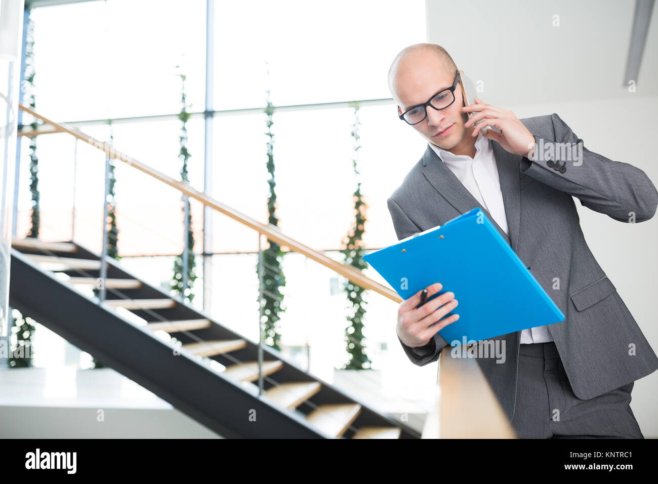Confident businessman holding clipboard while using smartphone by ...