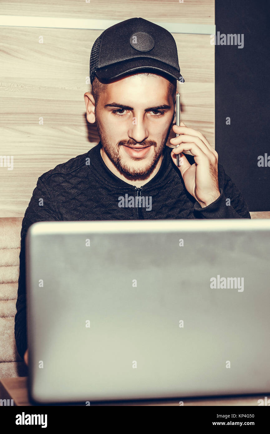 Young man with cap talking on the phone, using laptop computer and ...