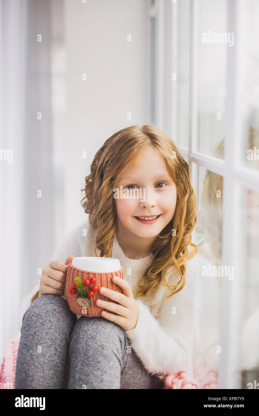 Portrait of cute happy little girl sitting on windowsill, drinking hot ...