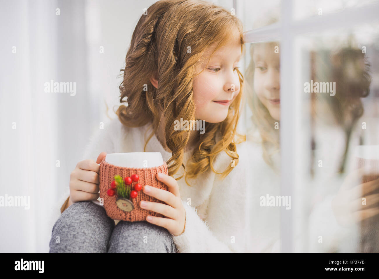 Portrait of cute happy little girl sitting on windowsill, drinking hot ...