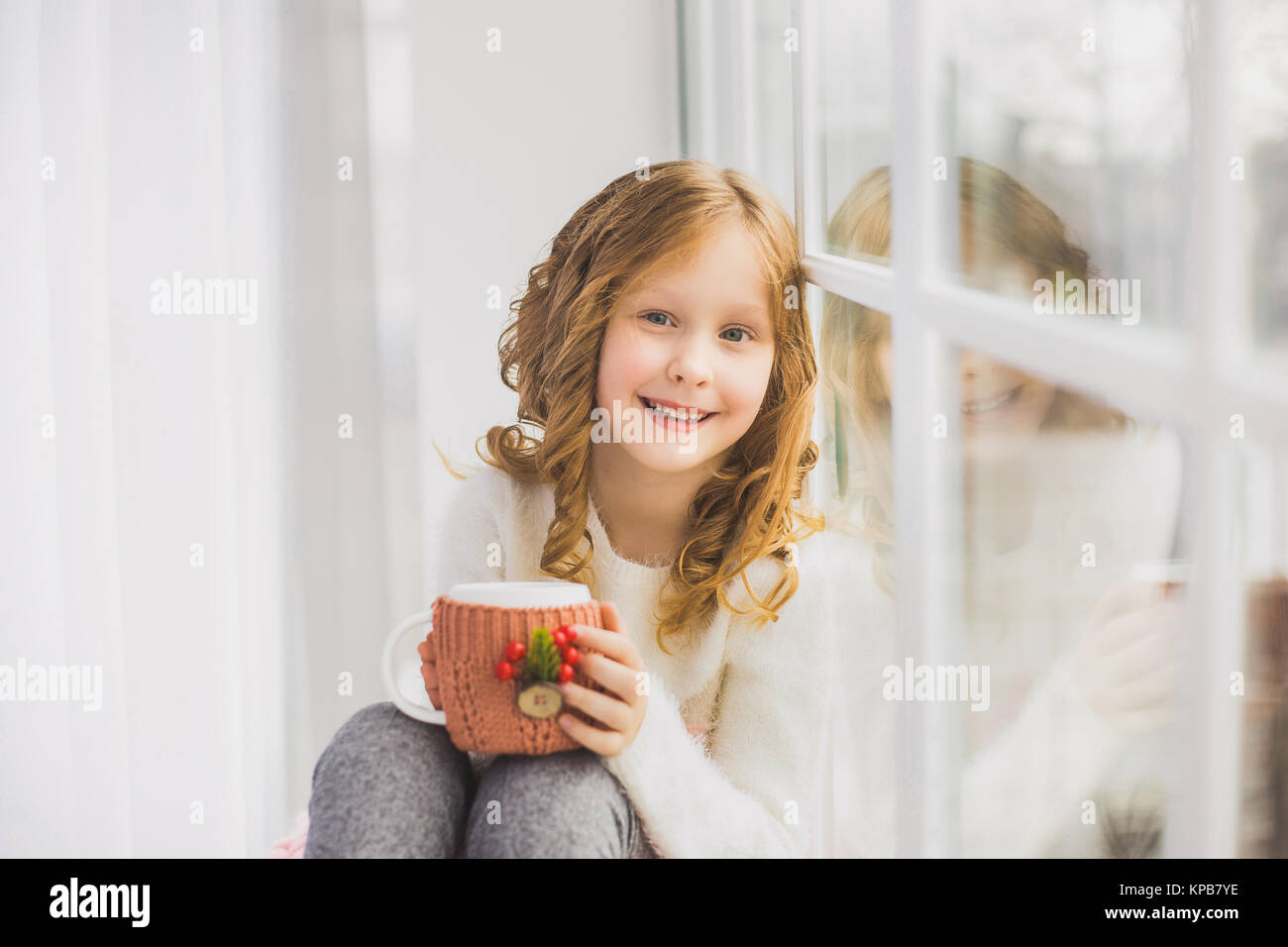 Portrait of cute happy little girl sitting on windowsill, drinking hot ...