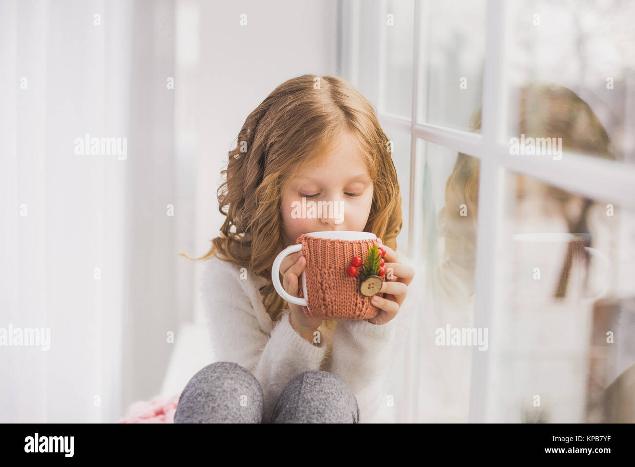 Portrait of cute happy little girl sitting on windowsill, drinking hot ...
