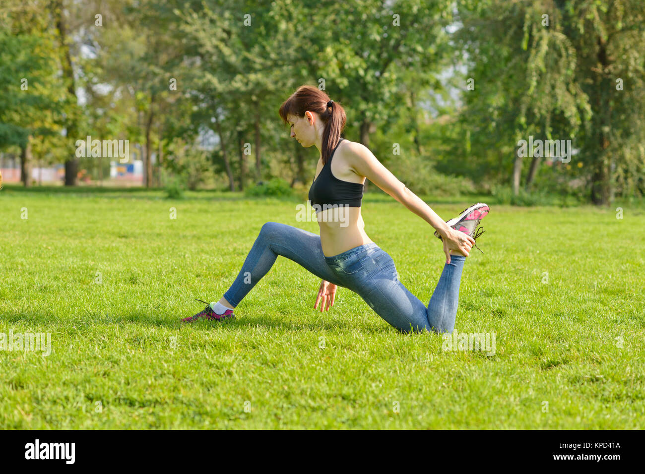 A girl is doing stretching exercises on a green grass in a public park ...