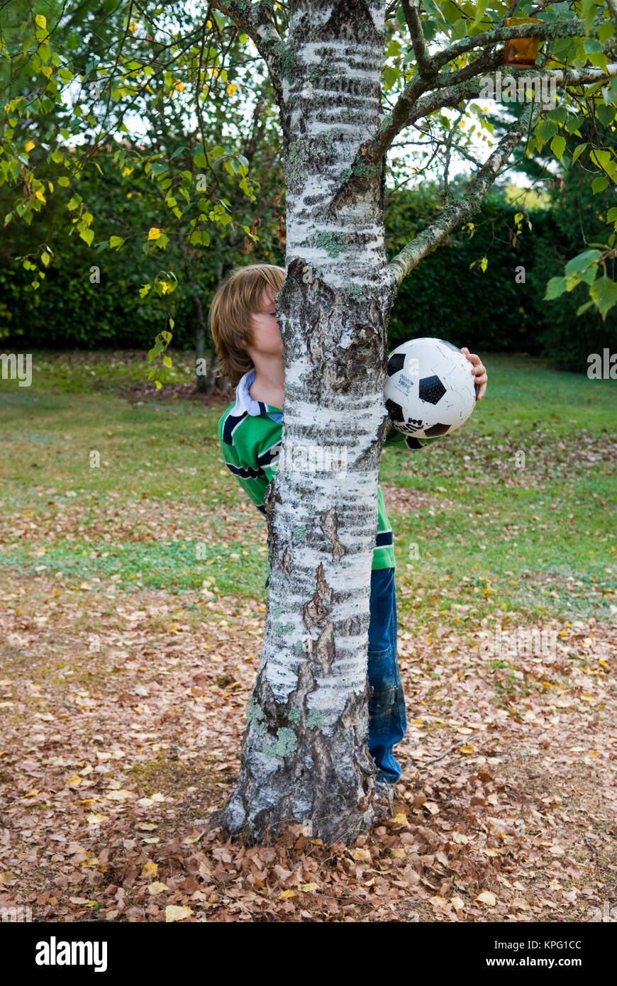 eight year old boy hides behind a tree in a private garden holding a ...