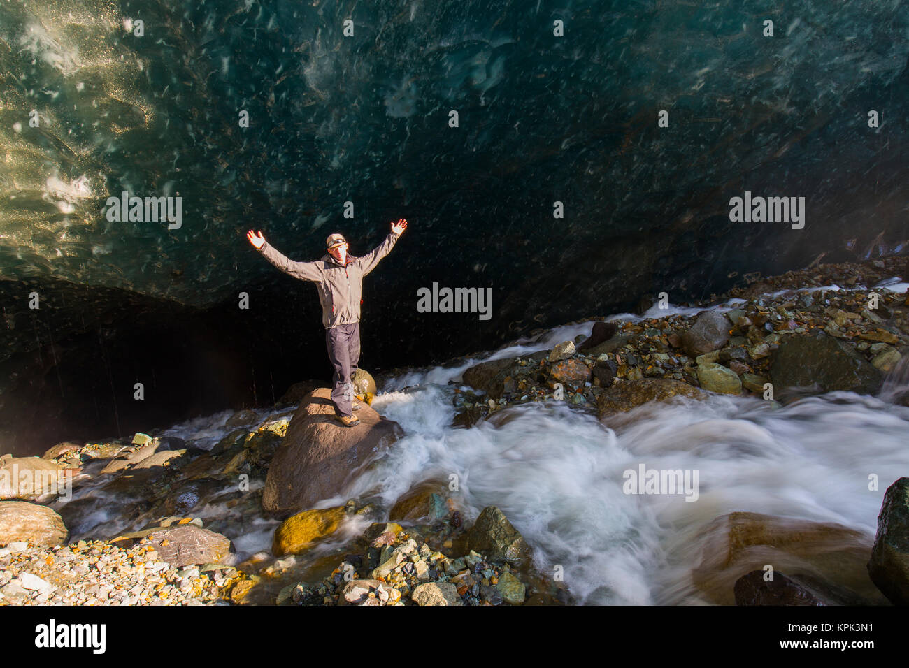 A man poses on a boulder with arms outstretched in front of a cave ...