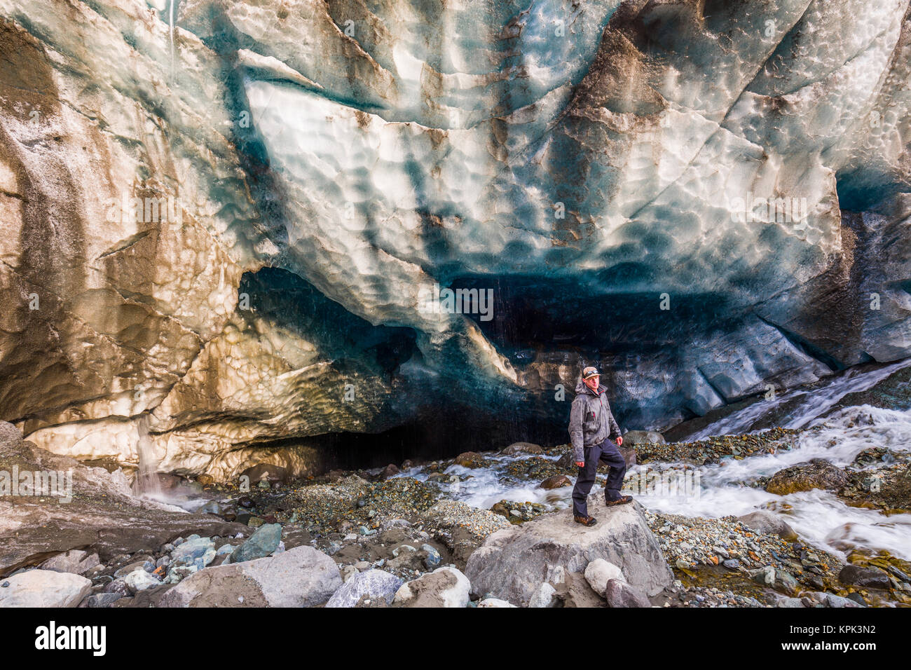 A man surrounded by rushing water poses in front of a cave beneath the ...