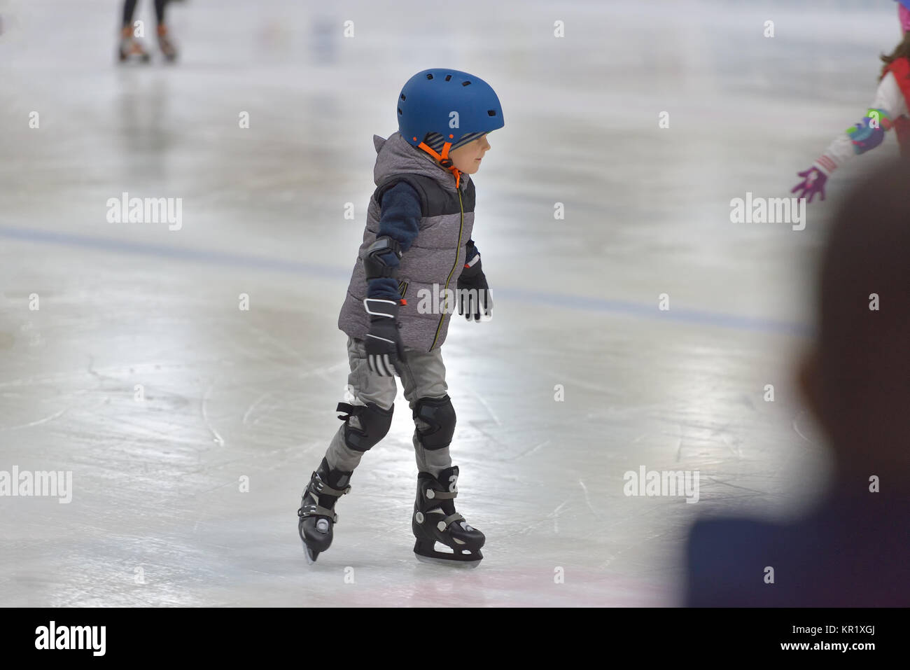 Adorable little boy in winter clothes with protections skating on ice ...