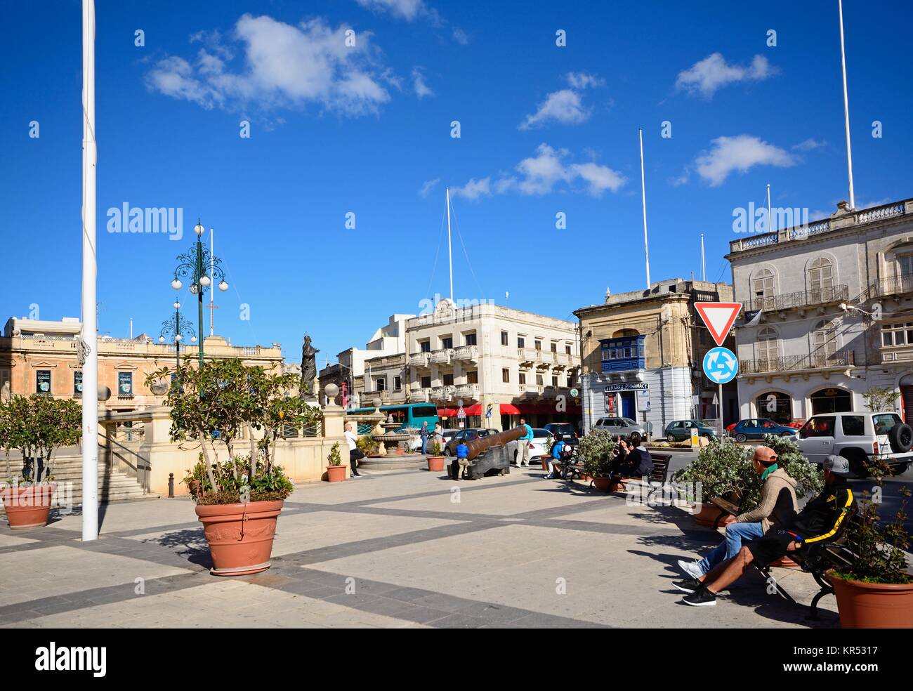 Tourists relaxing in the plaza by the Mosta Dome with buildings around ...