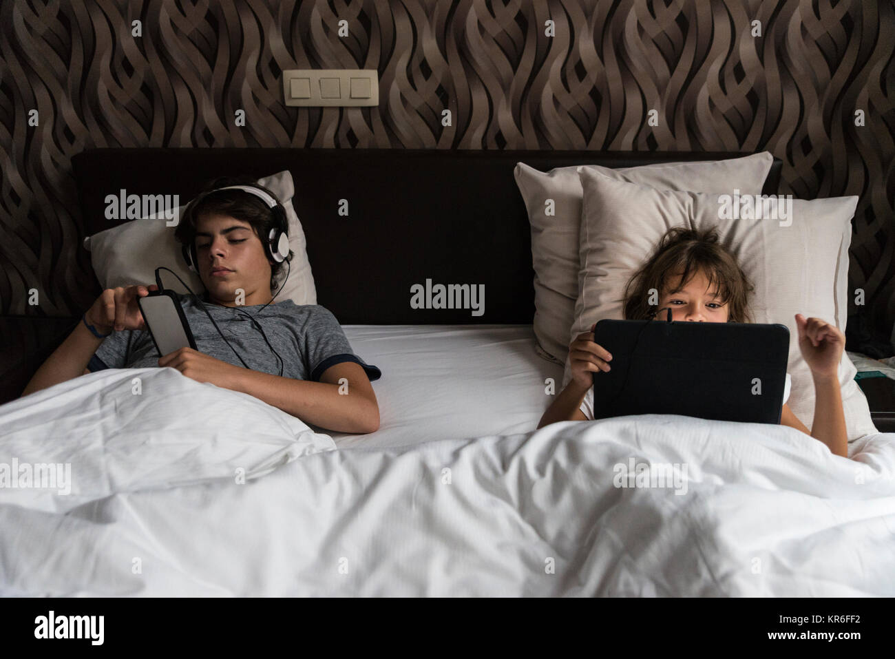 Boy and girl stretched out in bed relaxing and playing with smart phone ...