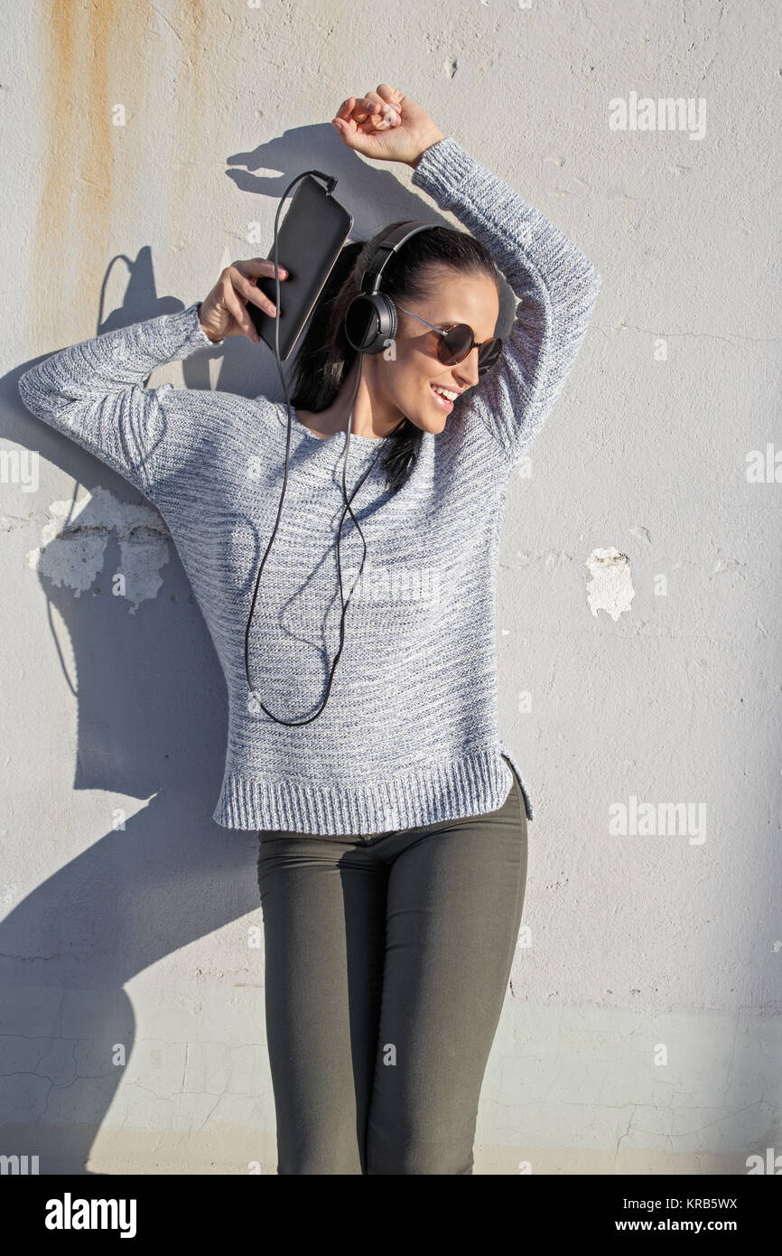 Young casual woman with headphones and tablet singing outdoor at wall ...