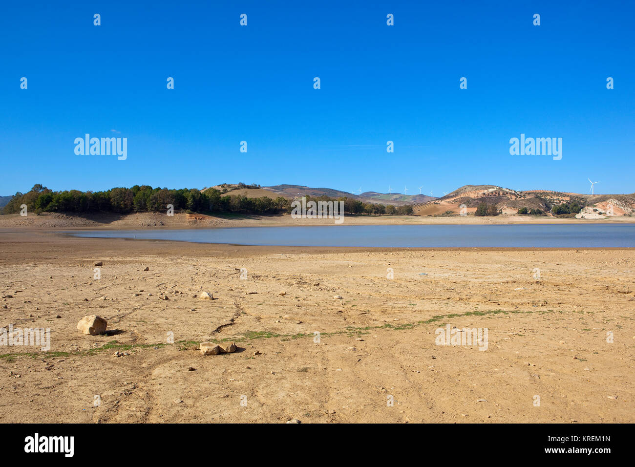 a dried up reservoir with sandy shores and rocks with woodland and wind ...