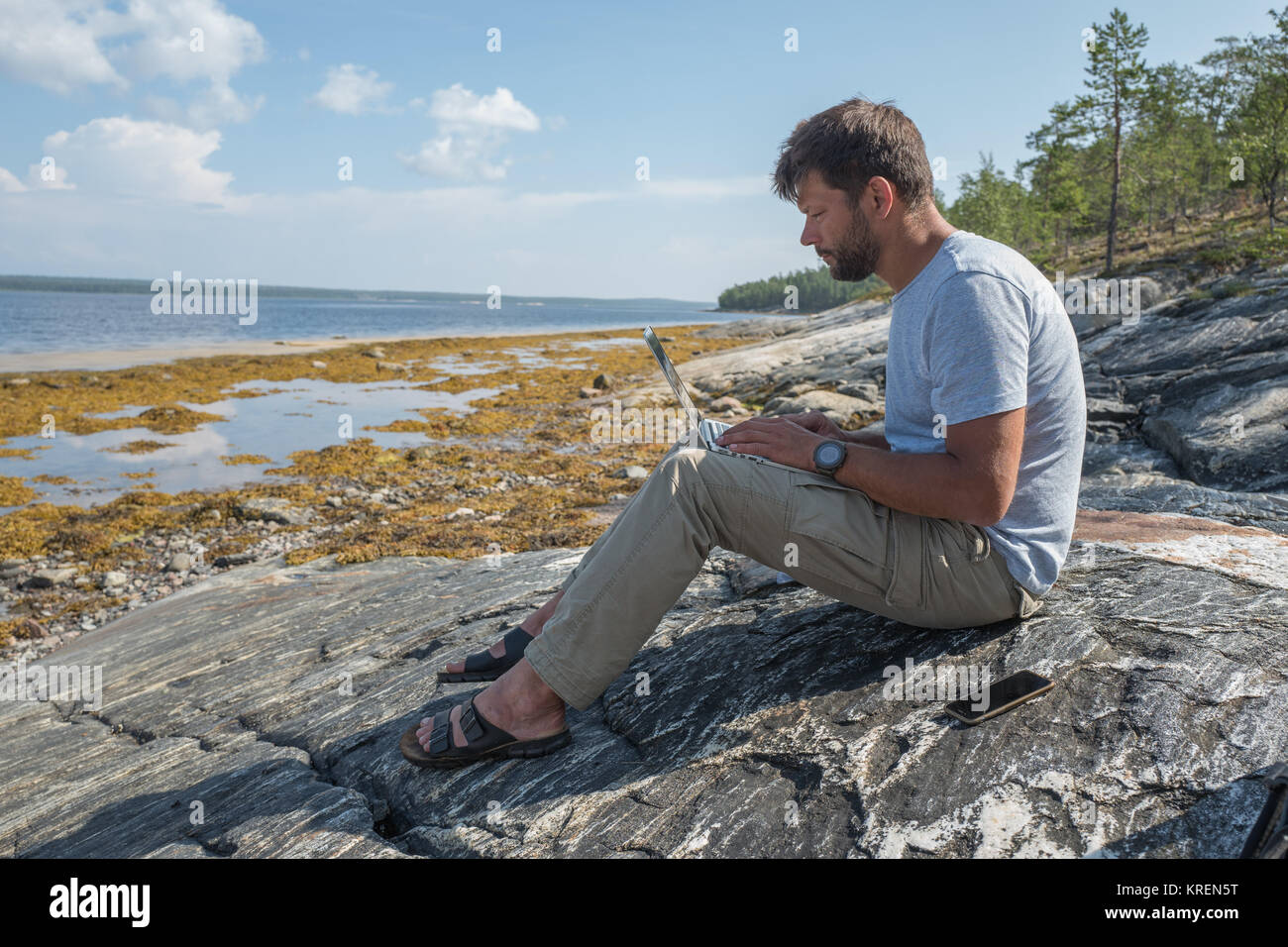 Handsome man is seating on the stone near of the sea and working with ...
