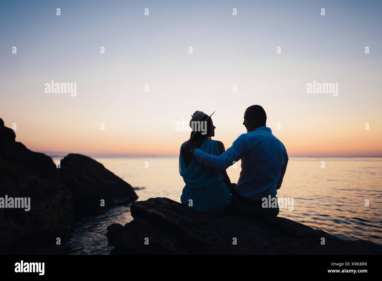 Silhouette of Caucasian couple sitting on rock near ocean at sunset ...