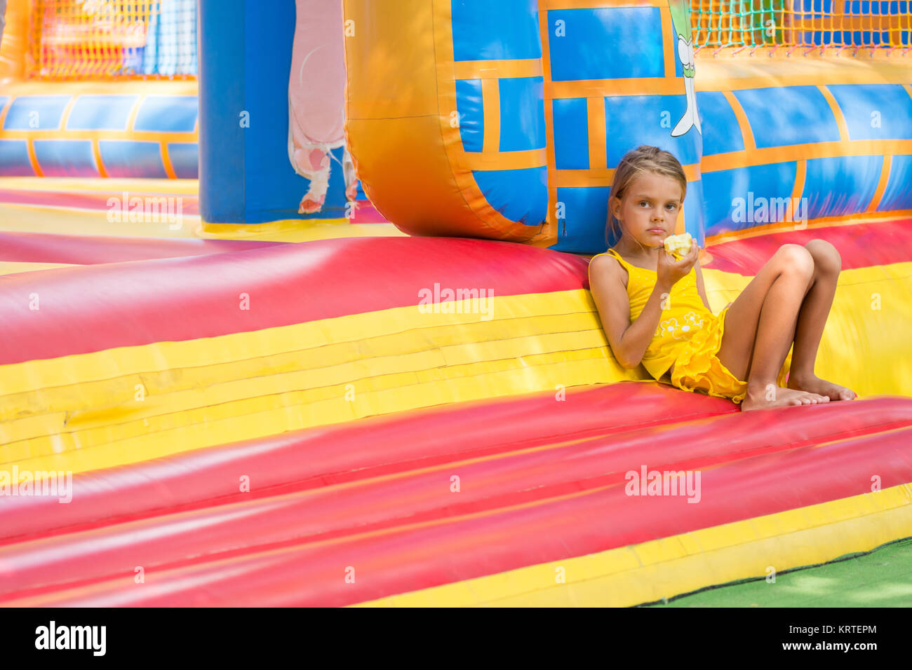 Girl sits at the entrance to an inflatable trampoline and eats an apple ...