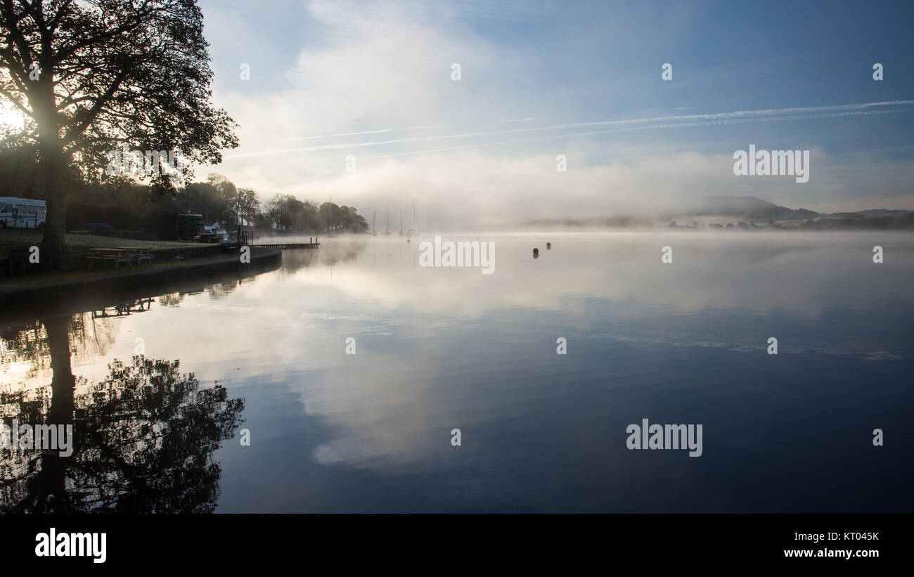 Mist rises from Windermere lake, beside woodland trees in autumn ...