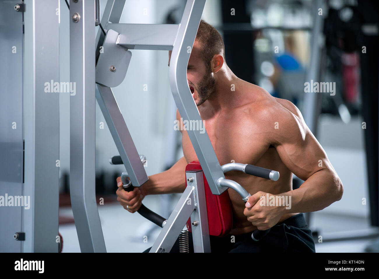 Determined young muscular man working on fitness machine at the gym ...