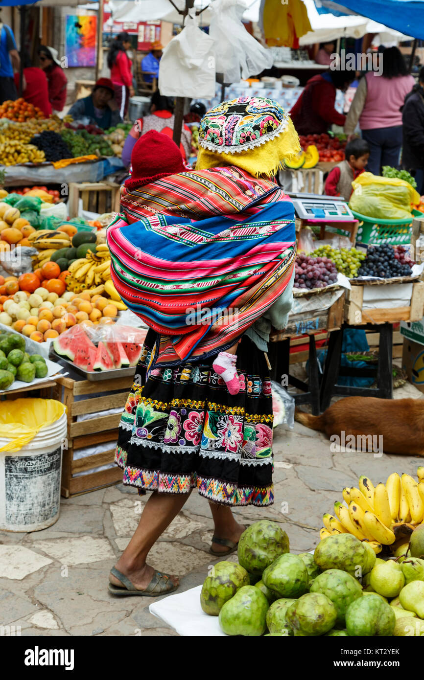 Quechua woman carrying baby on her back amid produce stalls, Pisac ...