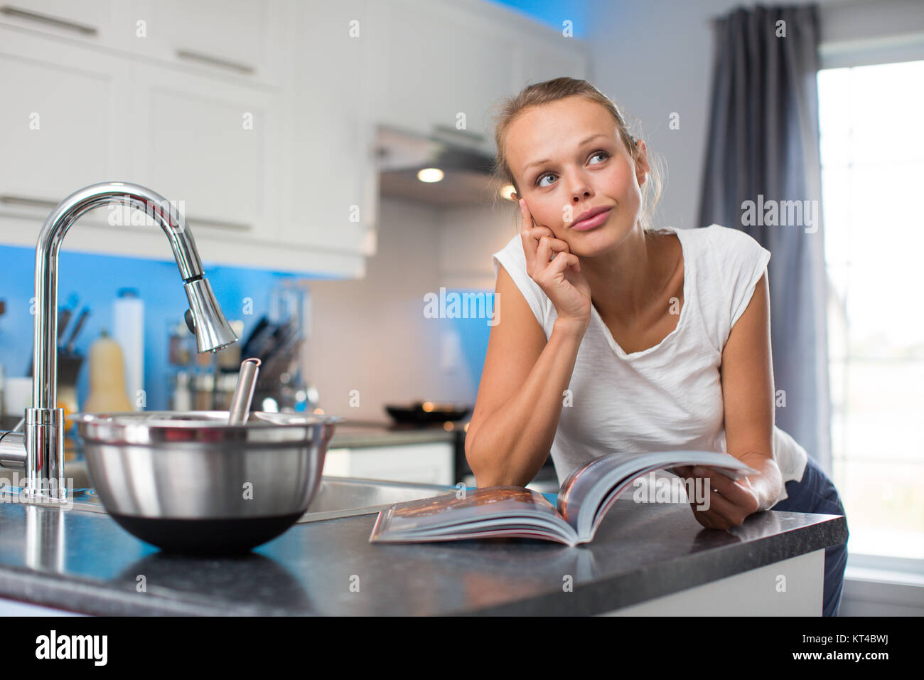 Pretty, young woman in her modern, clean and bright kitchen, thinking ...