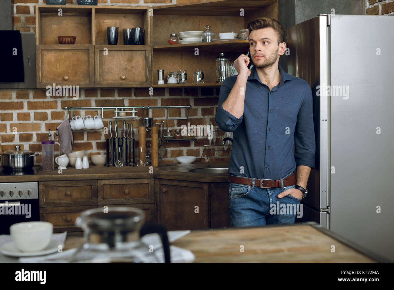Handsome young man leaning on refrigerator while talking on smartphone ...