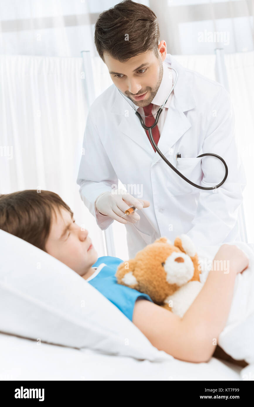 Scared little boy with teddy bear lying in hospital bed while doctor ...