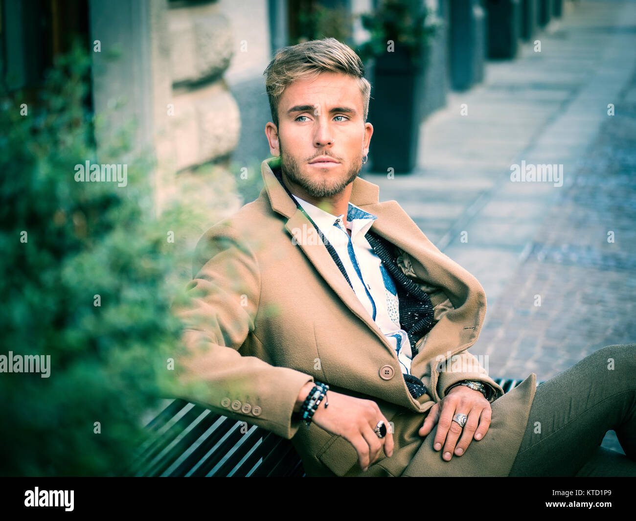 Handsome trendy blond man sitting outdoor in European city setting with ...