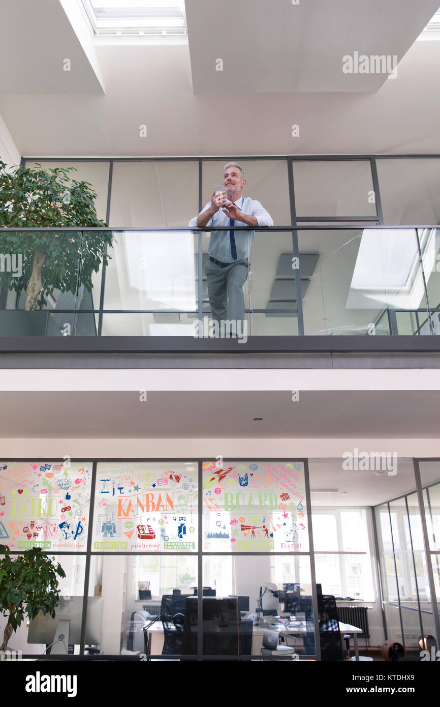 Successful businessman standing at railing in his office, using ...