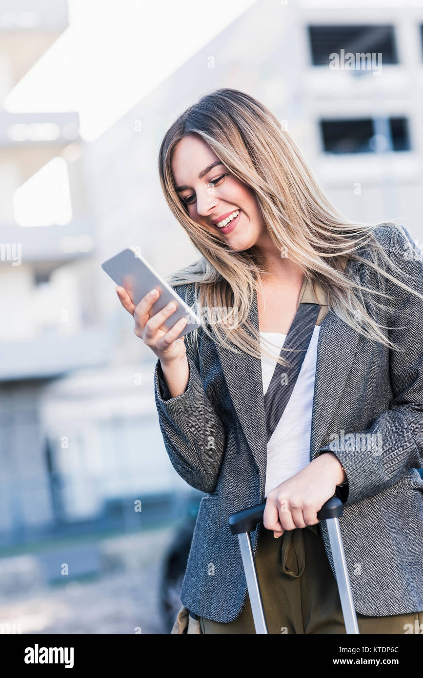 Happy young woman with rolling suitcase checking cell phone Stock Photo - Alamy