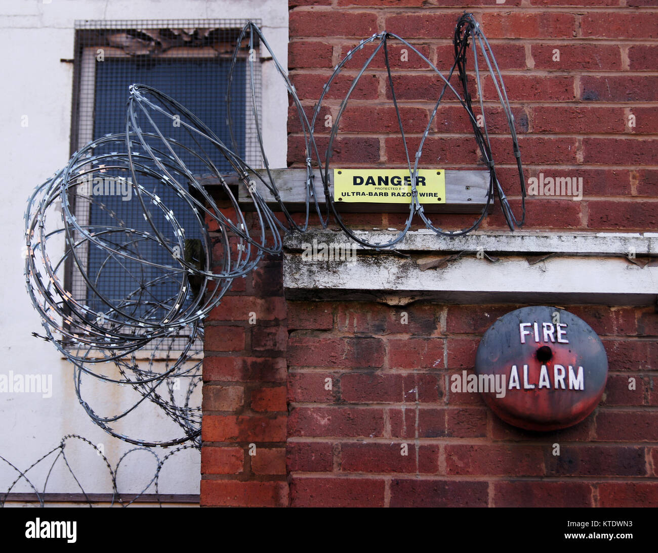 Razor wire against a white wall, with a rusty red fire alarm and yellow ...