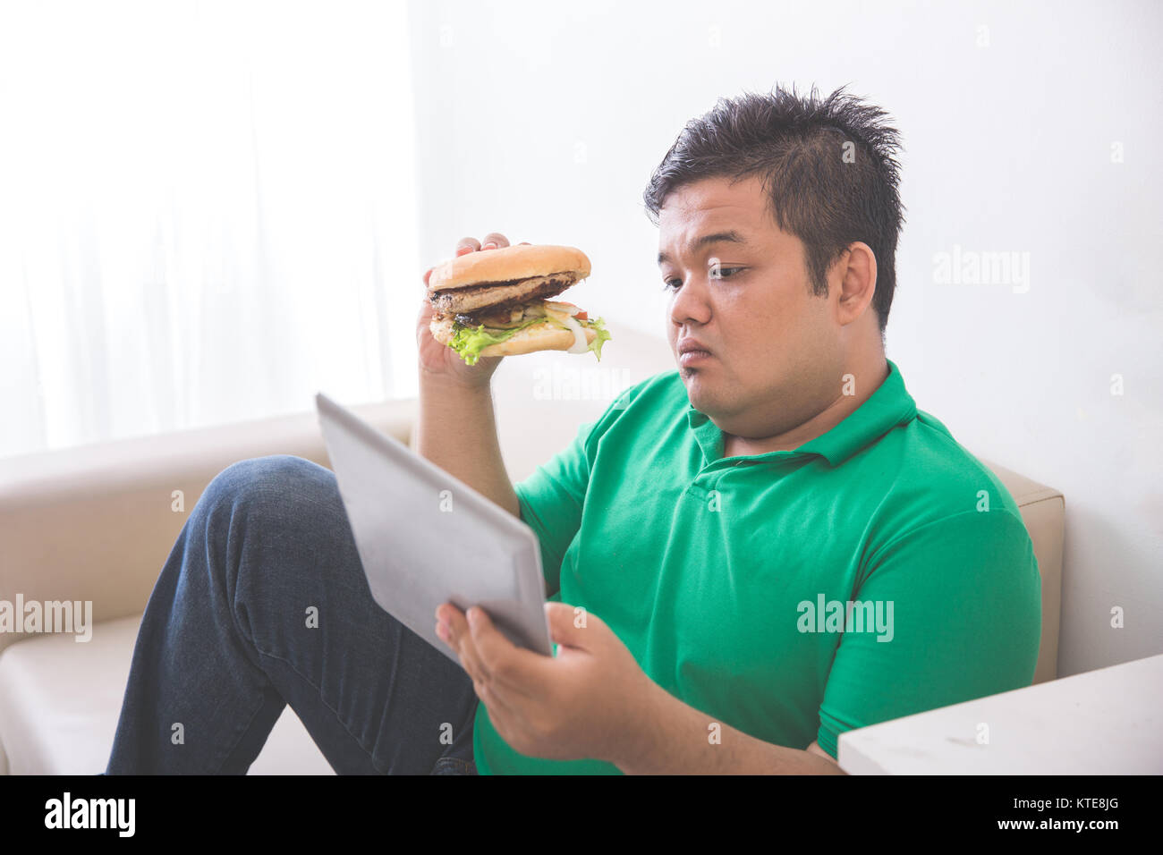 obese person eating hamburger while using tablet pc Stock Photo - Alamy