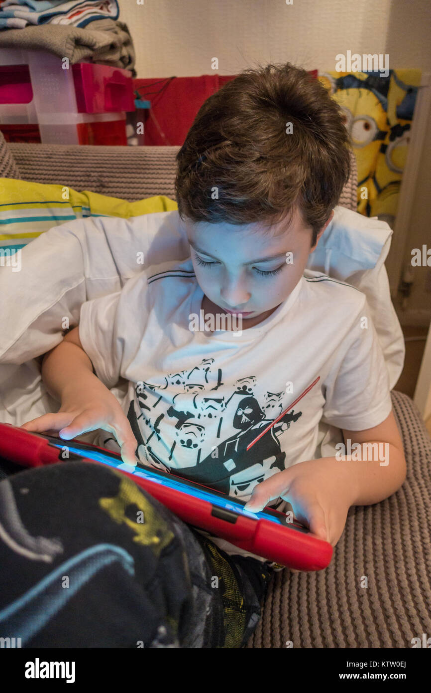 A young boy, dressed in pajamas, relaxes on a sofa playigng on his iPad ...