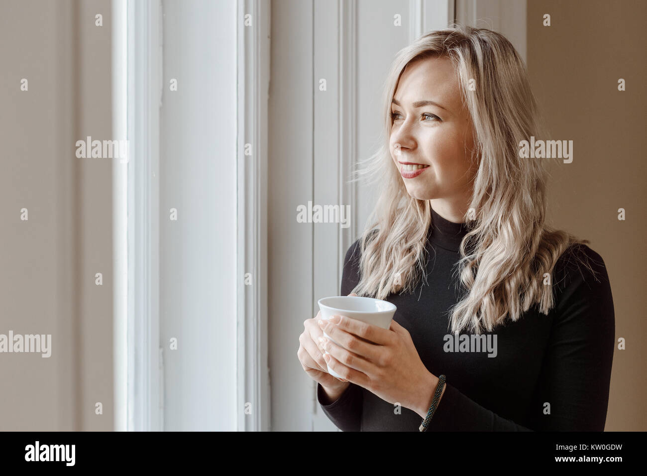 Beautiful young woman drinking tea or coffee standing by the window ...