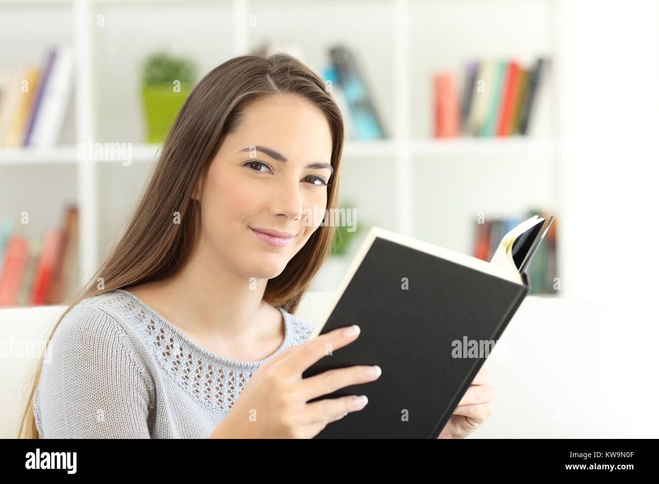 Lady reading a paper book and looking at you sitting on a sofa at home ...