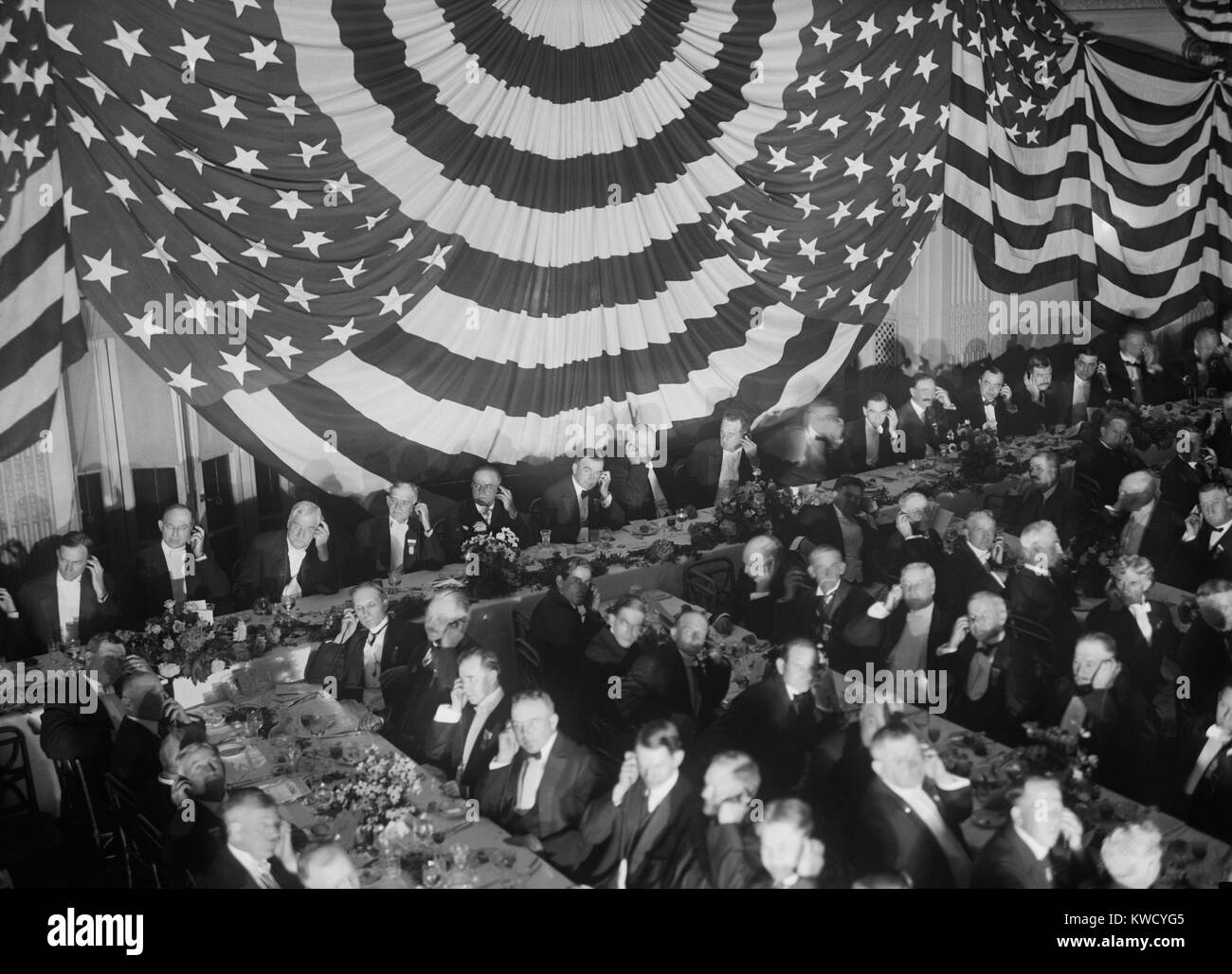 Banquet scene with seated men holding listening devices to their ears ...