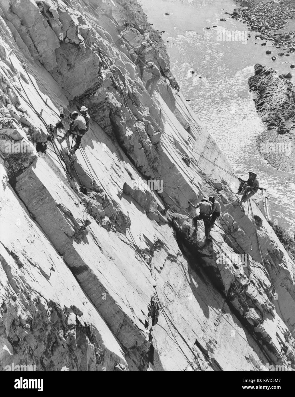 Eight men working high up on rock face of canyon wall, drilling holes ...