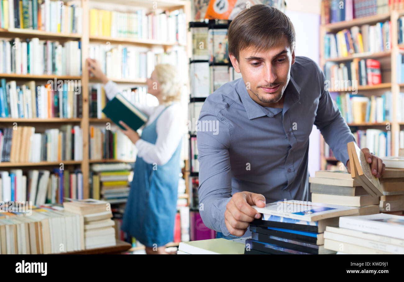 portrait of ordinary young man standing among bookshelves and searching ...