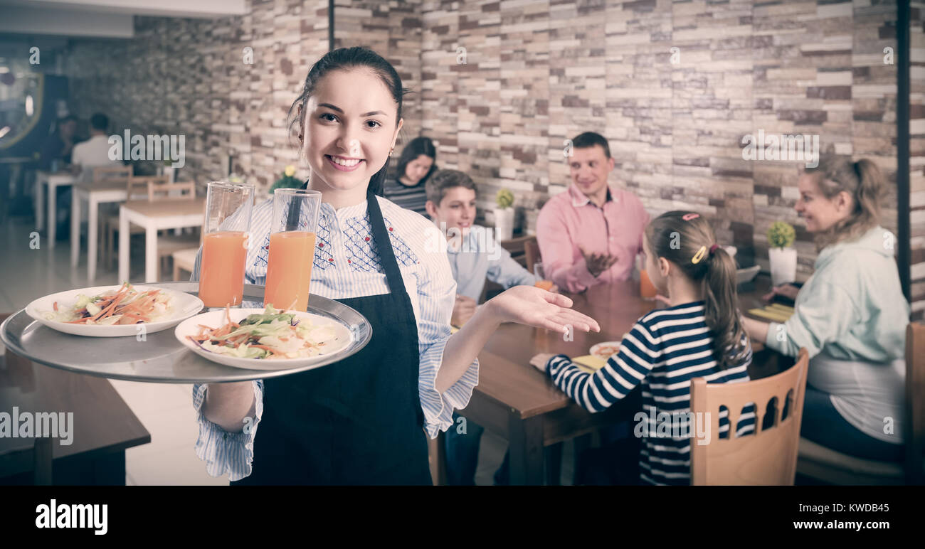 Cheerful waitress holding tray with dishes meeting visitors in cafe ...